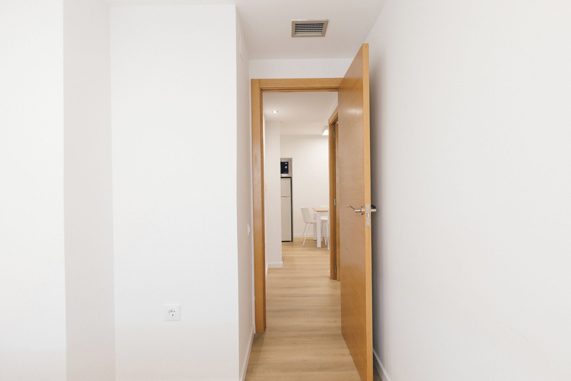Um corredor estreito com uma porta de madeira que dá para um quarto com uma mesa e cadeiras brancas.
