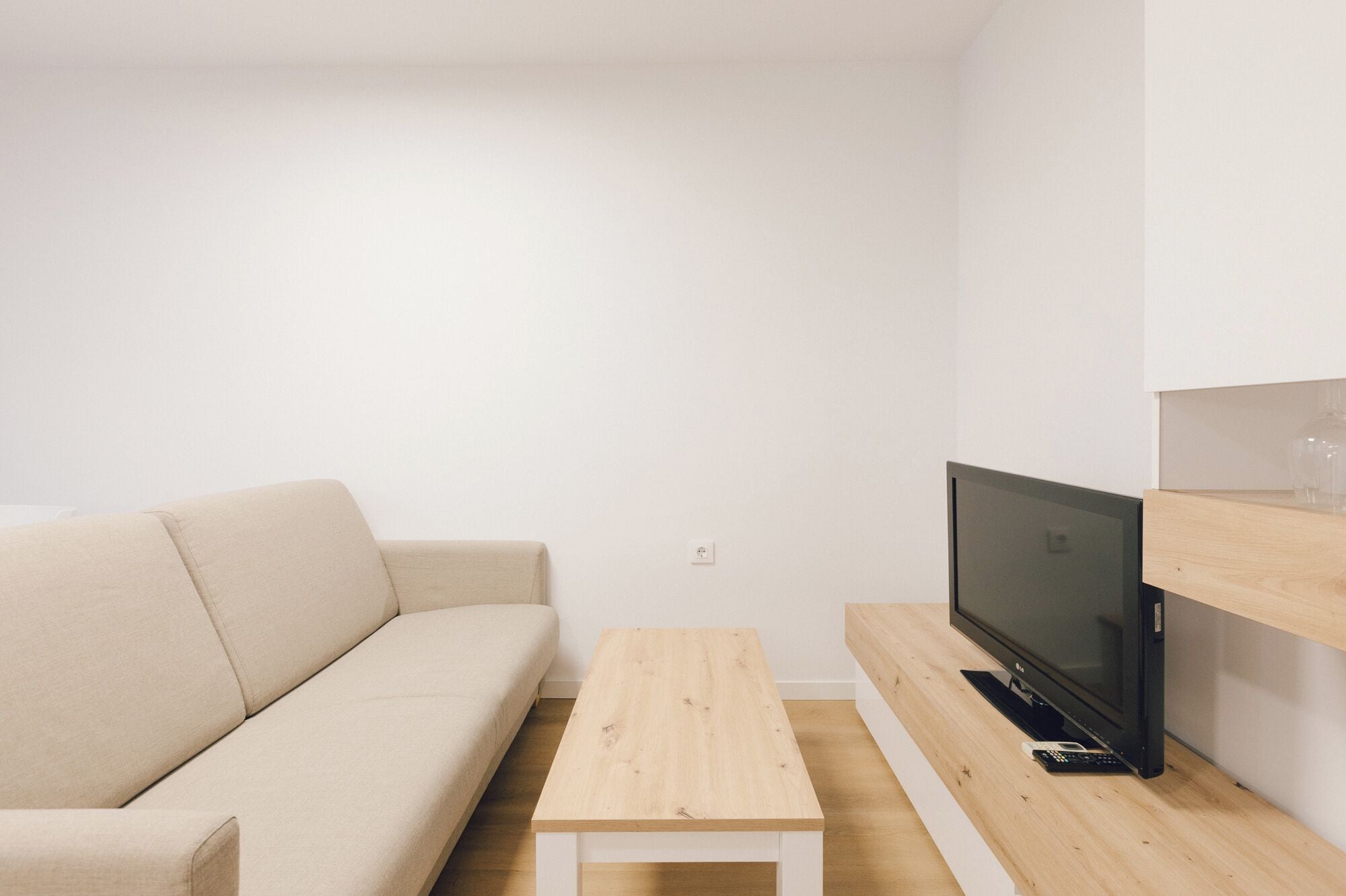 Uma sala de estar minimalista com um sofá bege, uma mesa de centro de madeira e uma televisão de tela plana sobre um suporte de madeira.