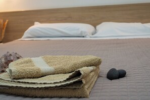 Cama bem arrumada com lençóis brancos, uma toalha bege dobrada e duas pedras escuras sobre a cama.