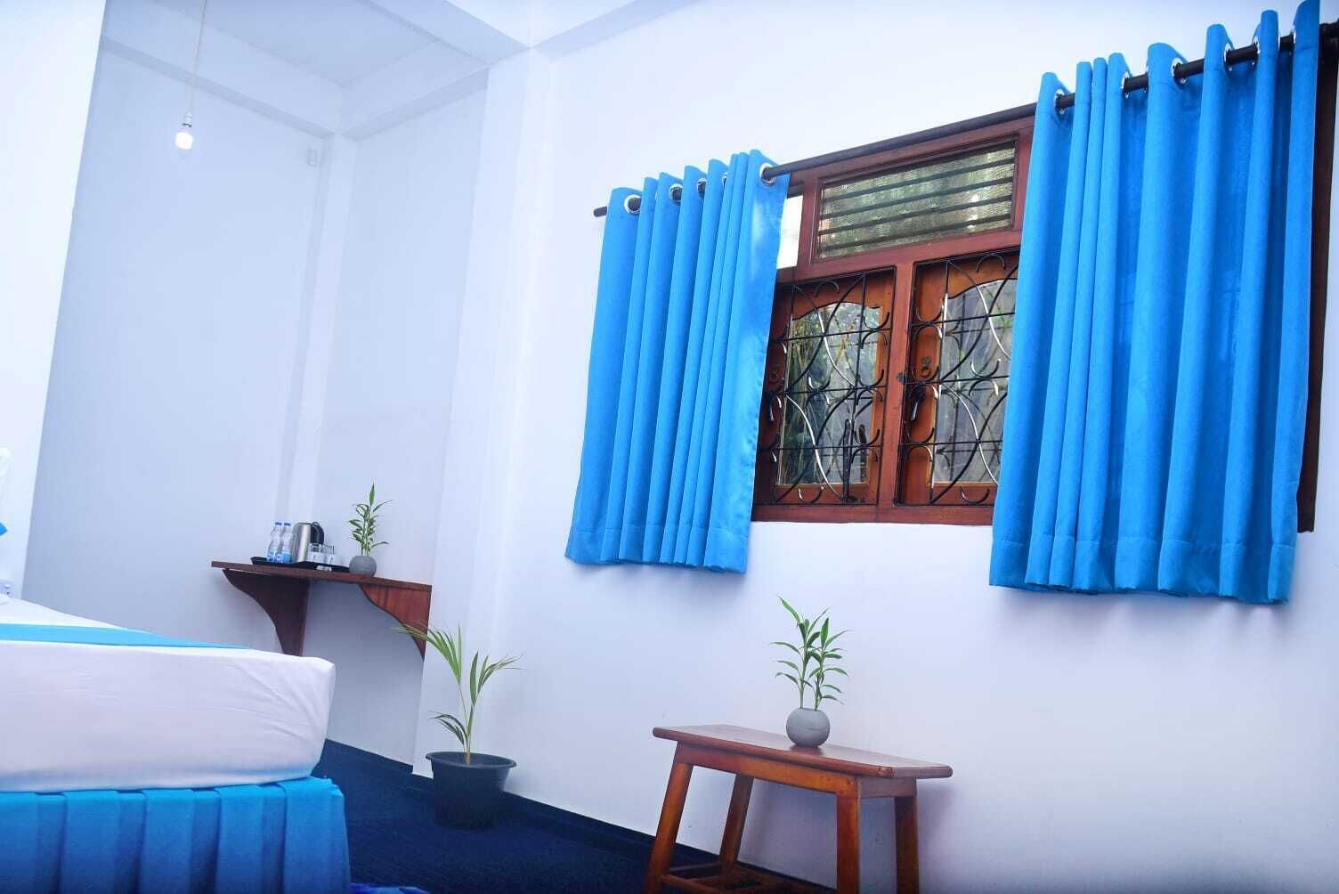 Um quarto com cama, janela com cortinas azuis, uma mesinha com planta em vaso e prateleira na parede com objetos.