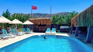 Uma piscina com espreguiçadeiras e guarda-sóis, uma bandeira e uma estrutura com telhado de palha.