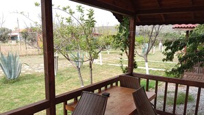 Alpendre coberto com mesa e cadeiras, com vista para um jardim com diversas plantas e um prédio ao longe.