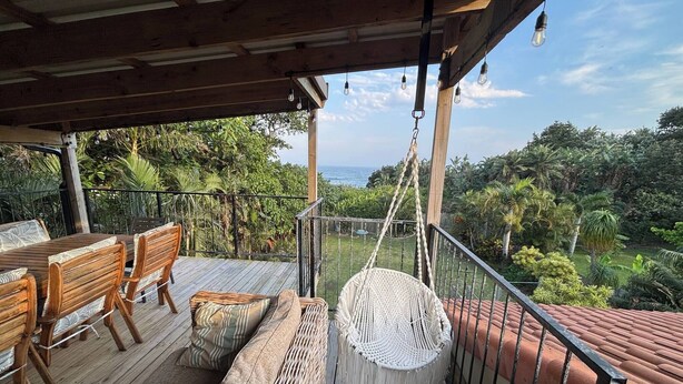 Um deck de madeira com mobiliário externo, uma cadeira de rede de macramé e vista para o oceano e vegetação.