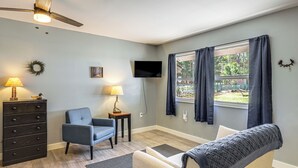 Uma sala de estar com ventilador de teto, TV fixada na parede, sofá, poltrona, cômoda e janela com cortinas.