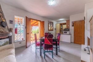 Uma sala de jantar com mesa e cadeiras, uma cozinha com eletrodomésticos, e uma vista através de uma porta aberta.