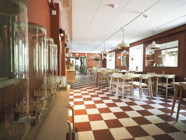 Interior de restaurante com chão quadriculado, mesas e cadeiras de madeira, luminárias pendentes e um balcão com copos.