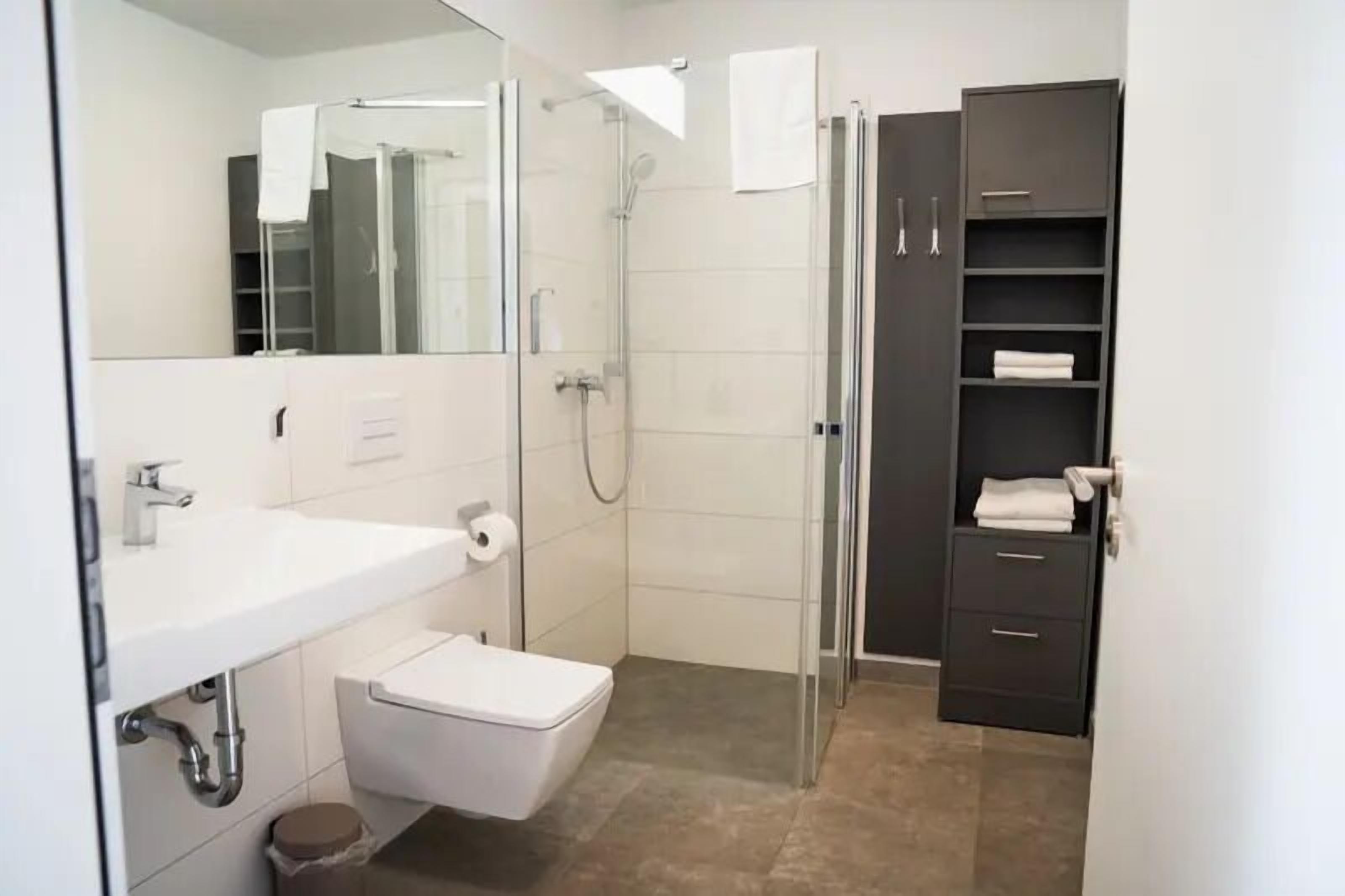 Uma casa de banho moderna com lavatório, sanita, chuveiro e um armário escuro com prateleiras.