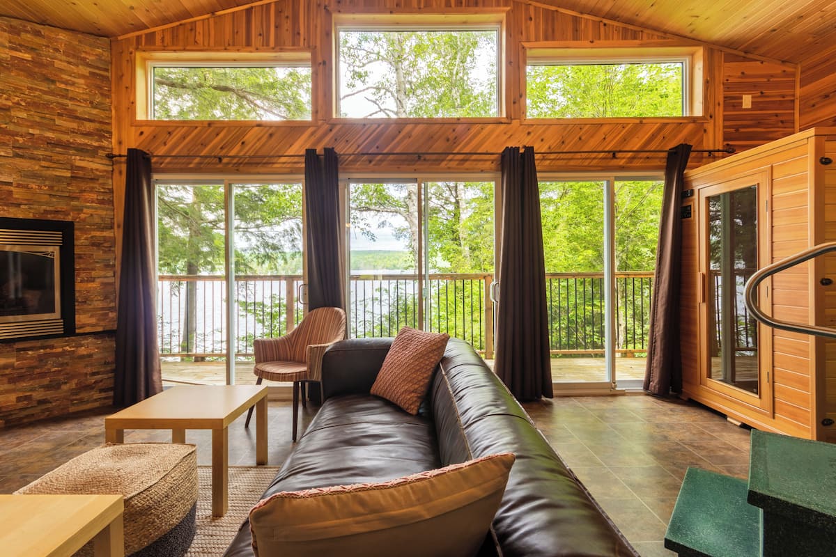 Interior de cabana com teto de madeira, sofá de couro, mesa e vista através de grandes janelas.