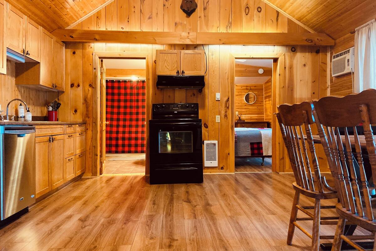 Interior de cabana de madeira com cozinha, área de jantar e uma cama visível ao fundo.