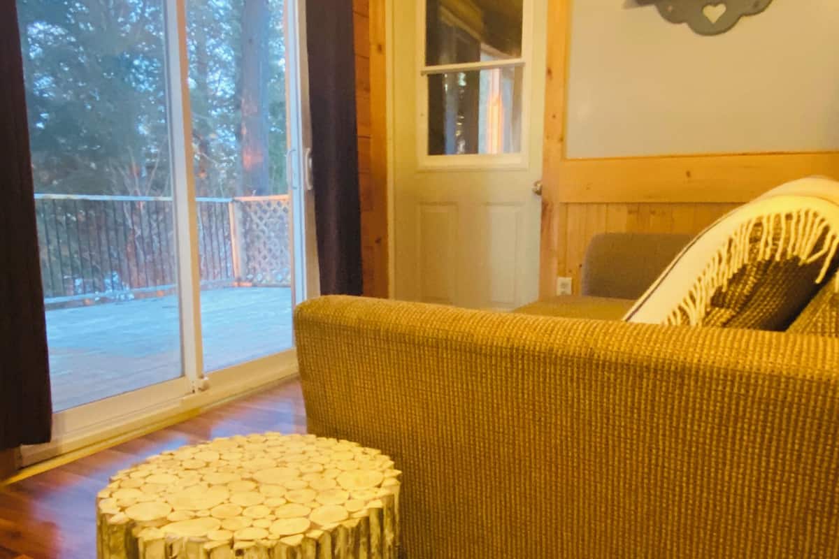 Quarto com piso de madeira, um banquinho de madeira redondo, um sofá amarelo, uma janela com vista para árvores e uma porta com uma prateleira acima.