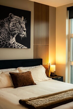 Quarto de hotel com uma grande obra de arte com padrão de leopardo na parede, uma cama com lençóis brancos, um travesseiro marrom e uma parede revestida de painéis de madeira, além de uma mesa de cabeceira com abajur.