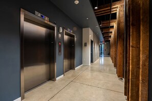 Corredor moderno com três elevadores, piso lustroso e lambris de madeira à direita.