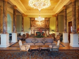 Um amplo hall com lareira, colunas ornamentadas e um candelabro.