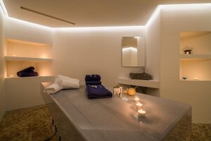 Sala de spa com mesa de massagem, toalhas, velas e prateleiras com objetos pessoais.