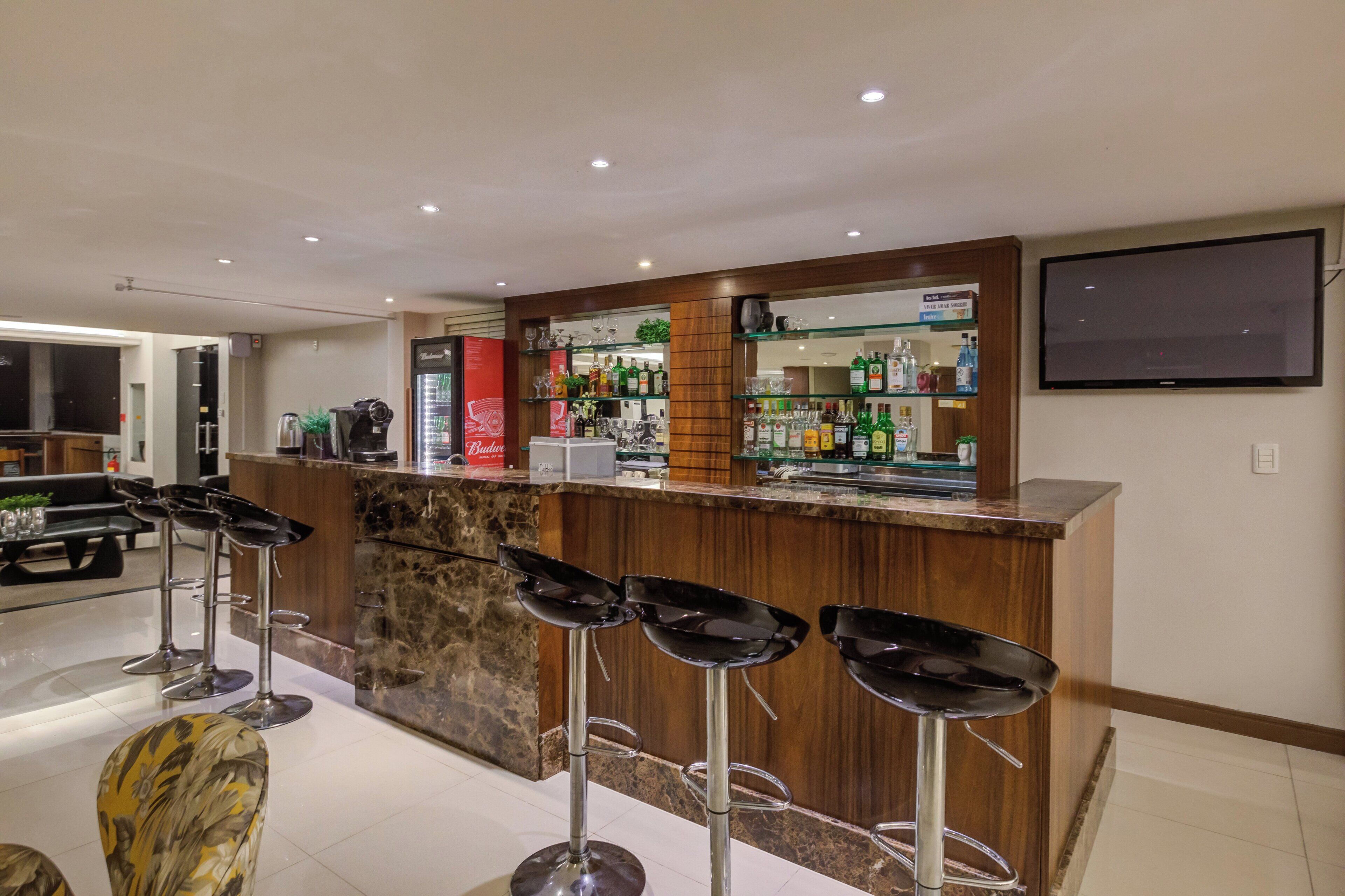 Uma área de bar com bancada de mármore, vários banquetes e uma variedade de garrafas de bebidas alcoólicas.