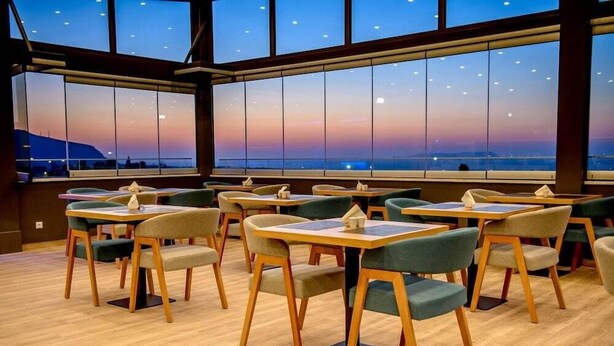 Um restaurante com várias mesas e cadeiras, amplas janelas com vista para o horizonte urbano ao entardecer.