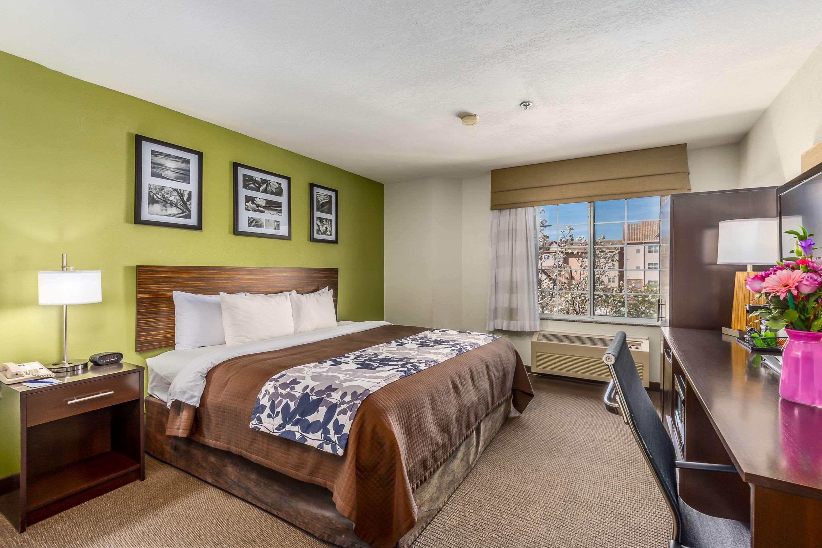 Quarto de hotel com uma cama grande, uma escrivaninha, uma cadeira, uma luminária e uma janela com vista para edifícios.
