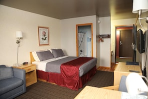 Quarto de hotel com cama, escrivaninha, cadeira, televisão, banheiro e relógio.