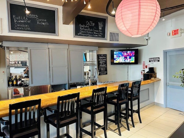 Uma área de bar com balcão de madeira, banquetas pretas, uma televisão na parede e um quadro negro com a lista de tipos de cerveja.