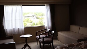 Quarto de hotel com sofá, mesa redonda pequena, cadeira e vista para um edifício através de uma janela com cortinas translúcidas.