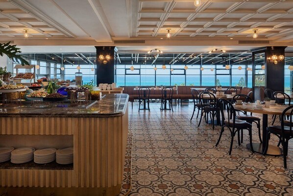 Uma sala de jantar espaçosa com vista para o mar, apresentando um bar central, várias mesas de jantar e piso estampado.