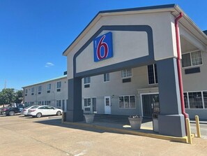 Um motel com um grande letreiro com o número 6, um estacionamento e a entrada do prédio.