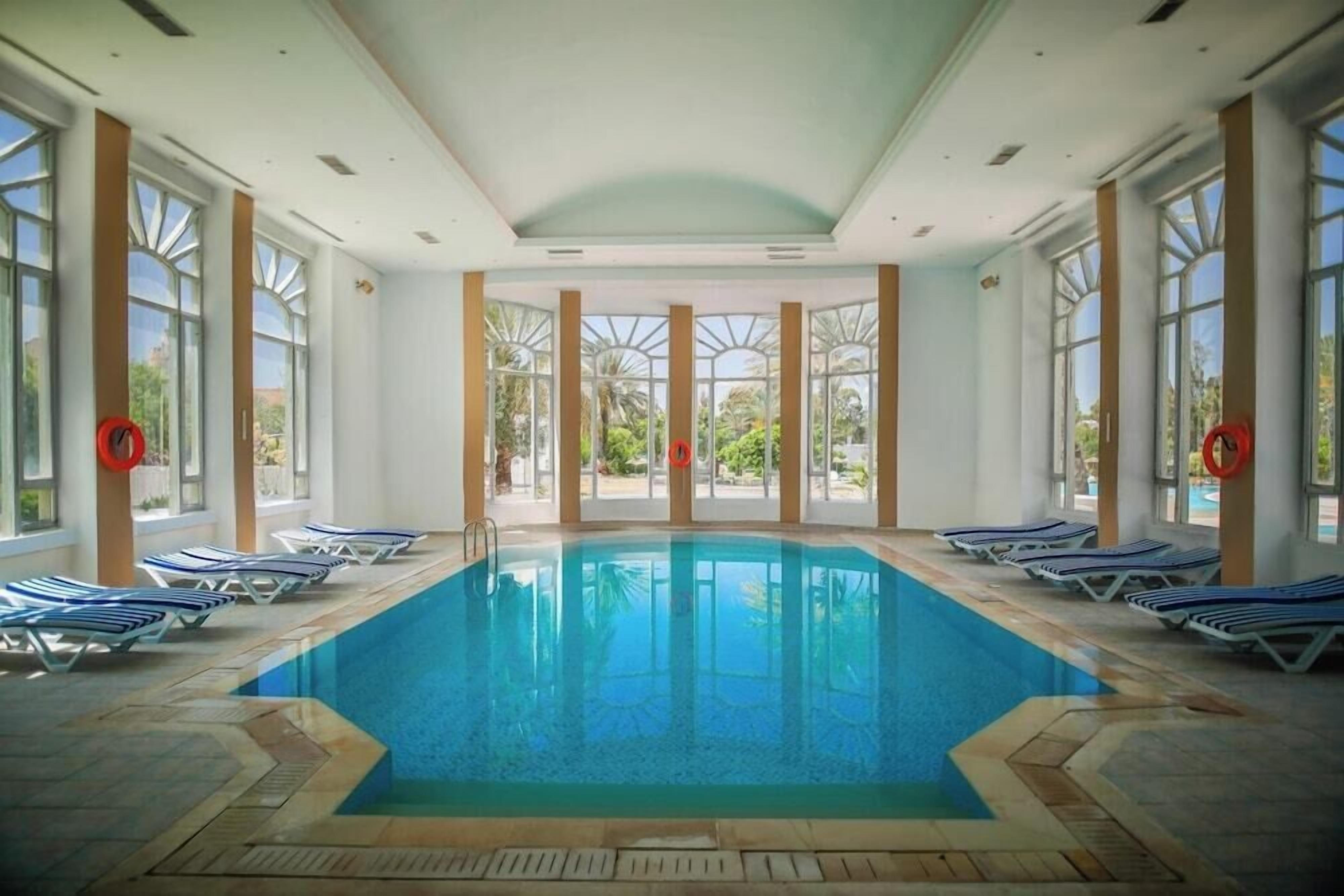 Piscina interior com paredes de vidro transparente, espreguiçadeiras e piso ladrilhado.