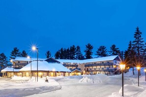 Um hotel com vários andares, fachada coberta de neve, rodeado por árvores e montes de neve.