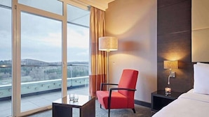 Quarto de hotel com uma janela grande, uma cadeira vermelha, uma mesa de cabeceira, um abajur e uma cama.