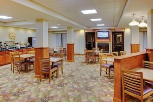 Uma sala de jantar de hotel com várias mesas e cadeiras, área de cozinha com eletrodomésticos e uma tela de televisão.