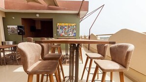 Um bar no terraço com mesas altas e cadeiras estofadas, uma pintura na parede e uma vista desobstruída da paisagem urbana.
