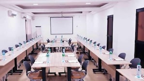 Uma sala de conferências com mesas compridas, cadeiras e uma tela de projeção.