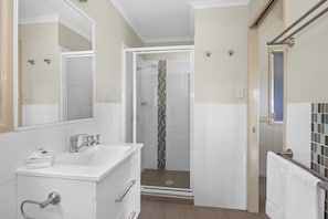 Casa de banho com lavatório branco, espelho, chuveiro com porta de vidro e um porta-toalhas.