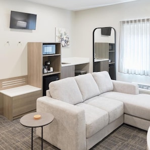 Quarto de hotel moderno com sofá, mesa redonda pequena, TV, máquina de café e espelho.