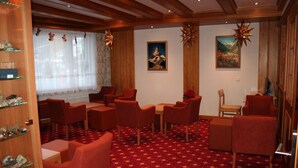 Quarto com painéis de madeira, carpete vermelho e cadeiras estofadas em vermelho. Há duas pinturas nas paredes, uma janela com cortinas leves e um enfeite em formato de estrela.