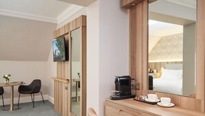 Quarto de hotel com escrivaninha, cadeiras, máquina de café e um espelho grande.