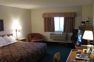 Quarto de hotel com cama, escrivaninha, computador, cadeira, abajur e janela com cortinas.