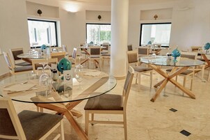 Uma área de refeições de hotel com mesas redondas de vidro, cadeiras de madeira e grandes janelas.