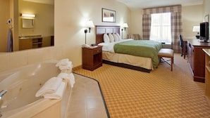 Quarto de hotel com uma banheira grande, uma cama com colcha verde, uma escrivaninha com computador e uma televisão.