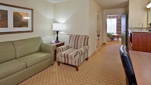 Quarto de hotel com um sofá verde, uma poltrona listrada, uma pequena mesa com abajur, uma escrivaninha de madeira e um banheiro visível por uma porta aberta.