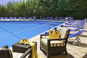 Uma área junto à piscina com espreguiçadeiras, uma cadeira de vime e uma bolsa amarela, além de uma mesa com frutas.