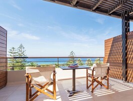 Uma varanda com mesa e duas cadeiras, com vista para uma praia com árvores e o oceano.