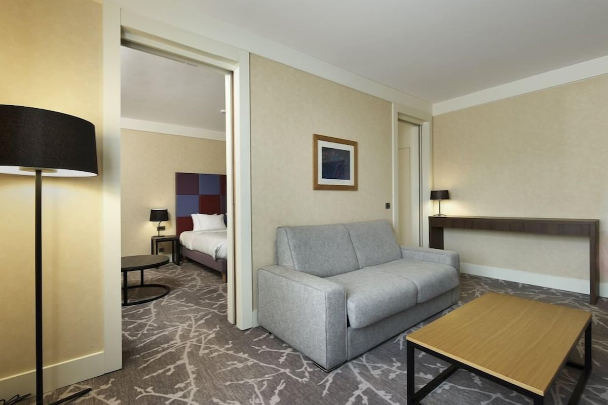 Quarto de hotel com sofá cinzento, mesa de centro de madeira, cama com cabeceira, pequena mesa com abajur e quadro na parede.