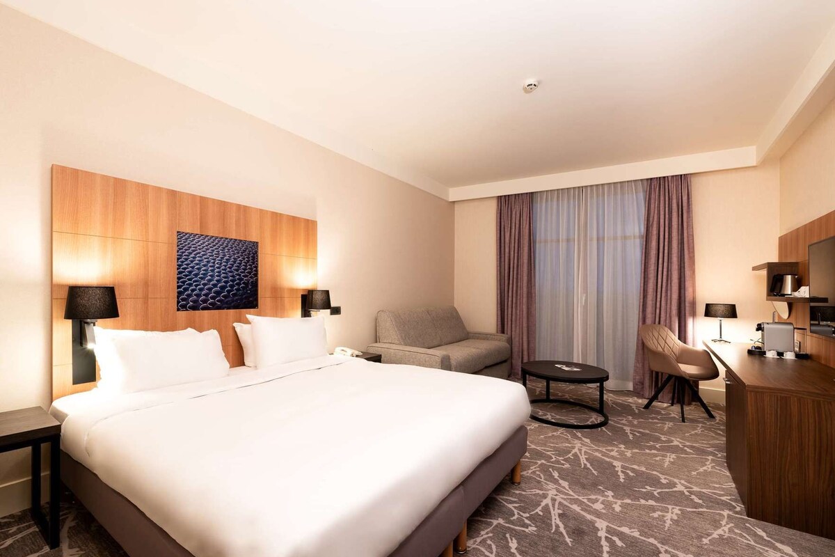 Quarto de hotel com uma cama grande, uma escrivaninha, uma cadeira e uma mesinha.