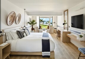 Quarto de hotel moderno com uma cama grande, televisão, escrivaninha e vista para a praia.