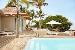 Uma área à beira da piscina com espreguiçadeiras, um deck de madeira e uma estrutura com telhado de palha.