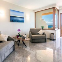 Uma sala de estar moderna com sofá, poltrona e mesa de centro. Há um quadro na parede, um vaso de planta e uma mala no chão.