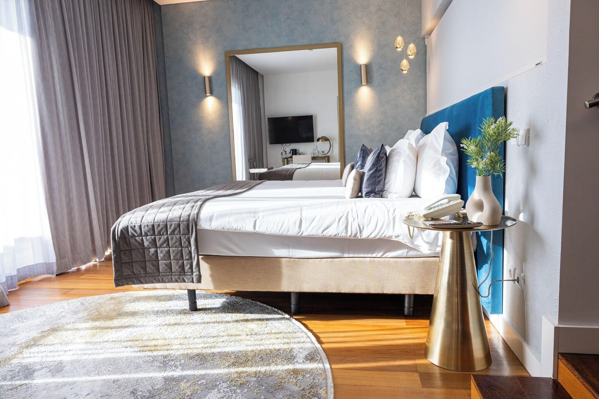 Quarto de hotel moderno com uma cama grande, mesa de cabeceira com telefone, espelho e uma parede azul de destaque.