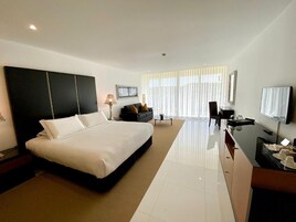 Quarto de hotel moderno com uma cama grande, uma escrivaninha, televisão e uma área de estar com sofá e poltronas.