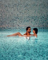 Um casal deitado em uma piscina azul cristalina, com um revestimento de azulejos estampados.